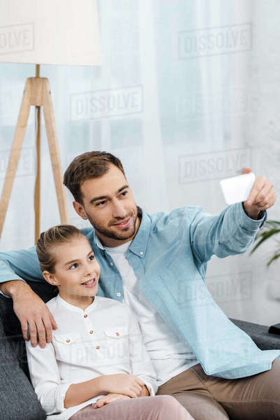 Smiling Father And Daughter Sitting On Sofa And Taking Selfie In Living