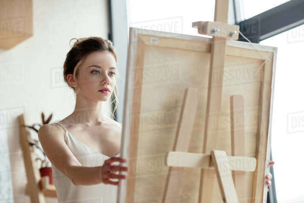 Beautiful young artist looking at picture on easel - Stock Photo - Dissolve