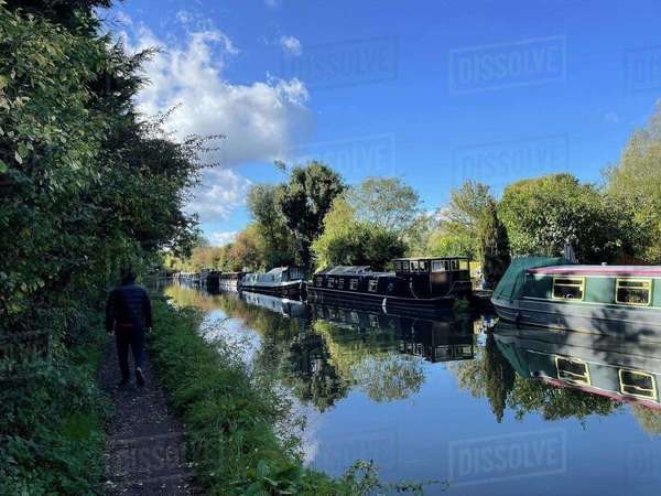 Rear view of a man walking along a towpath and Narrowboats moored along Grand Union Canal ...