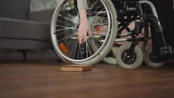 Woman moves wheelchair trying to reach book lying on floor. Female person with disability ...