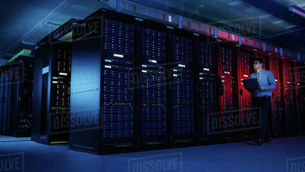 In Data Center: Male IT Specialist Walks along the Row of Operational Server Racks, Uses Laptop ...
