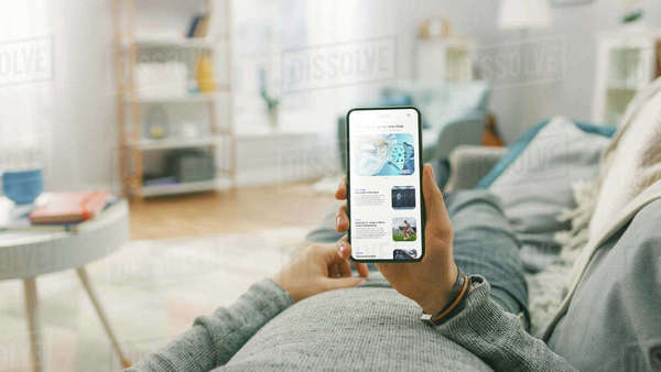 Young Man at Home is Lying on a Sofa and Using Smartphone for Scrolling and Reading News about ...