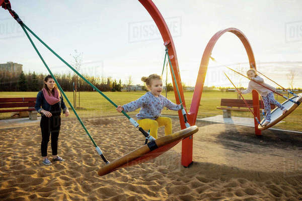 A young mom and her daughters playing on saucer swings in a playground on a warm autumn evening ...
