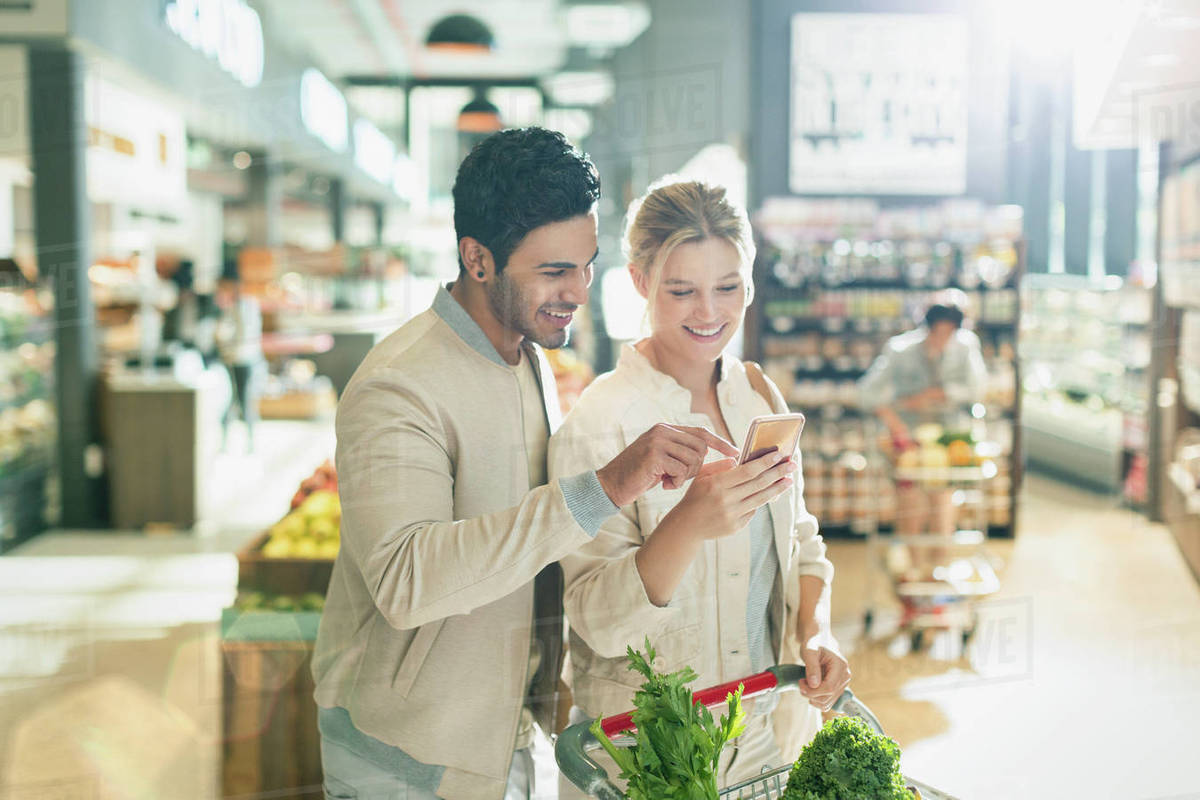 Young couple using cell phone, grocery shopping in grocery store market ...