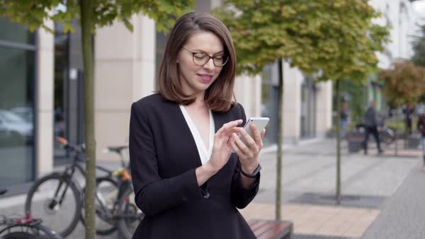 Smiling modern brunette in trendy formal blazer and glasses texting via ...