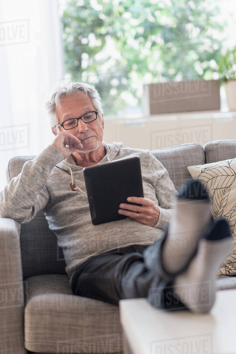 Senior man sitting on couch in living room and using tablet pc ...