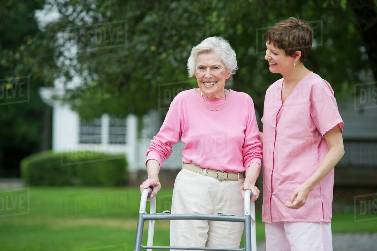 Senior woman walking with walker with help of nursing assistant - Stock ...
