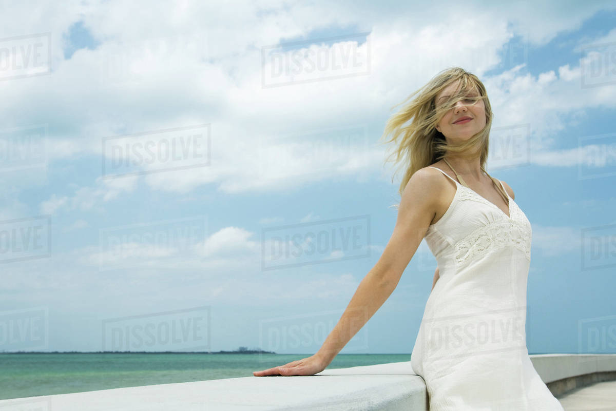 Young woman leaning against ledge with eyes closed, wind blowing hair ...