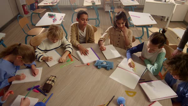 Children studying at round table in class. Teacher talking with ...