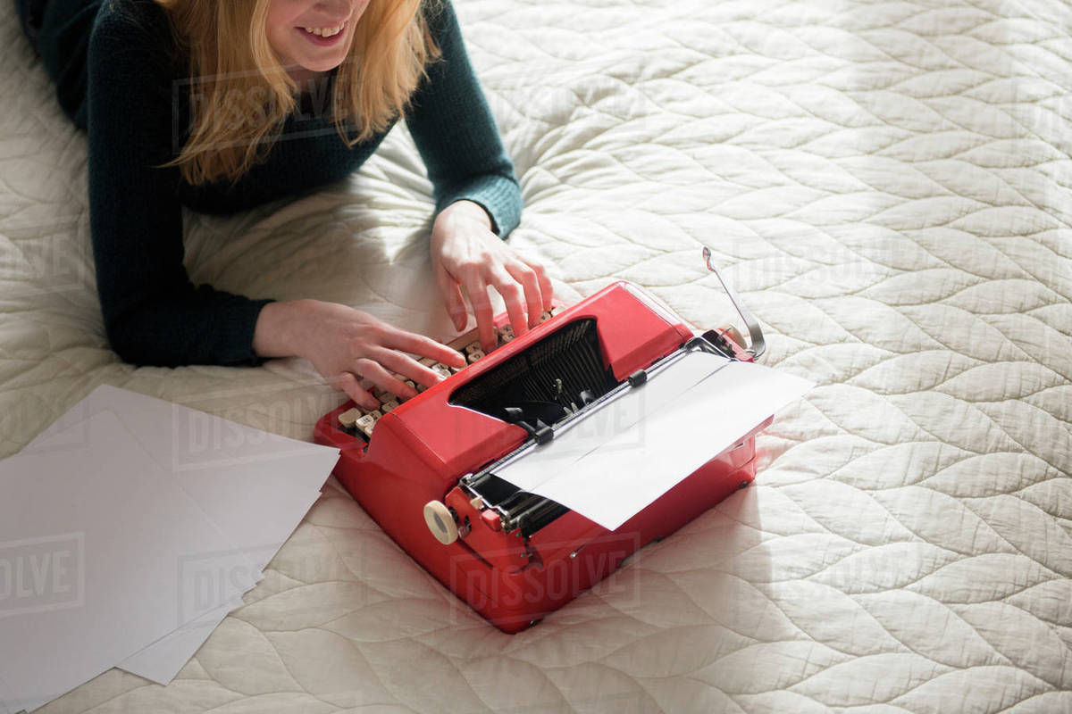 Smiling woman laying on bed typing on typewriter - Royalty-free Stock ...