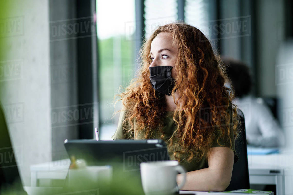Thoughtful young woman with face mask back at work in office after ...