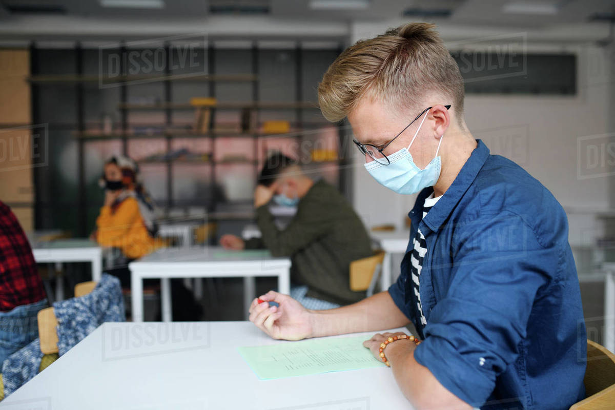 Side view of young student with face masks at desks at college or ...