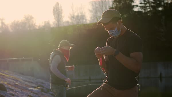 Wide shot of a man tying artificial fly to fishing rod string while his ...