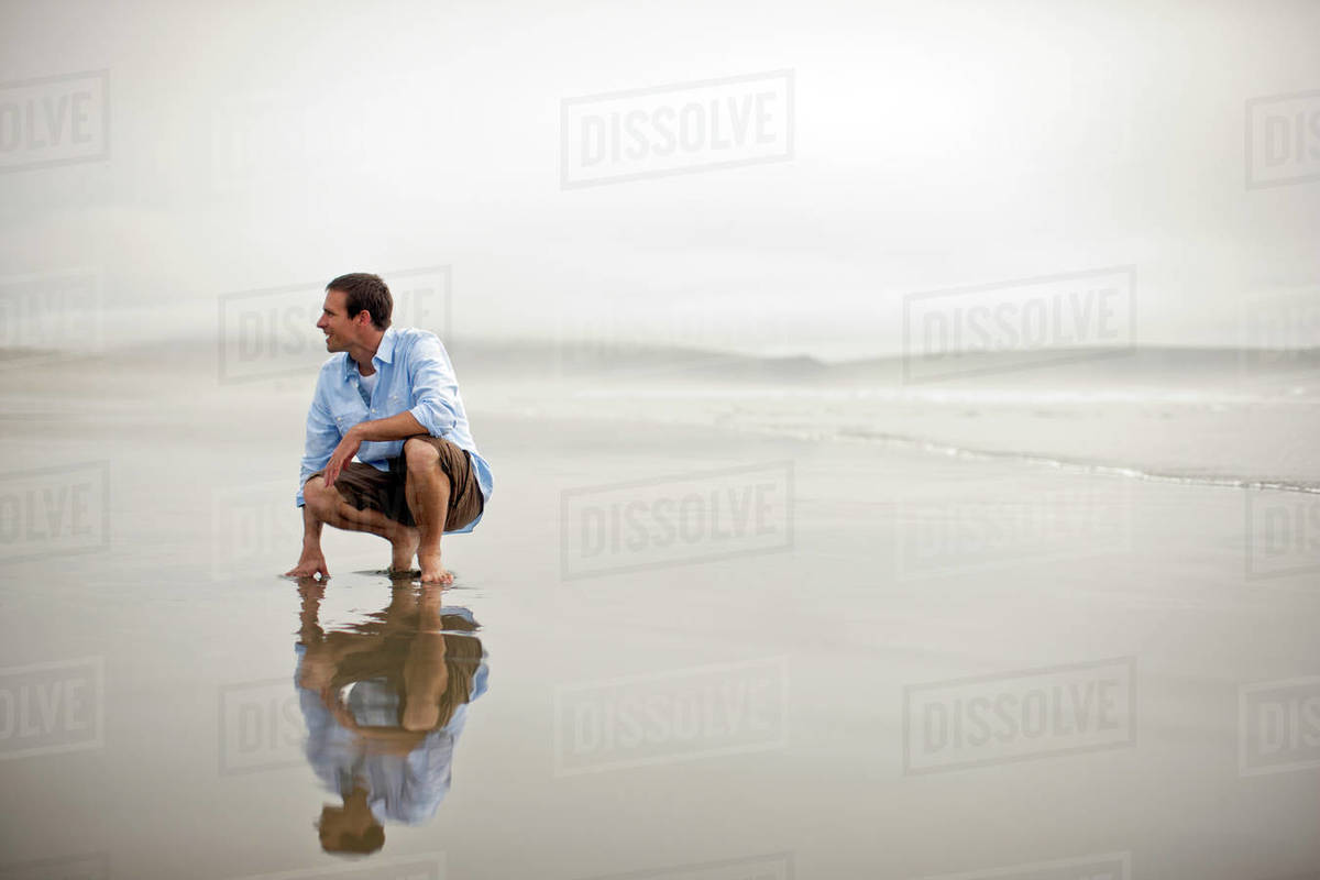 Cheerful man crouching on a beach at low tide. - Stock Photo - Dissolve