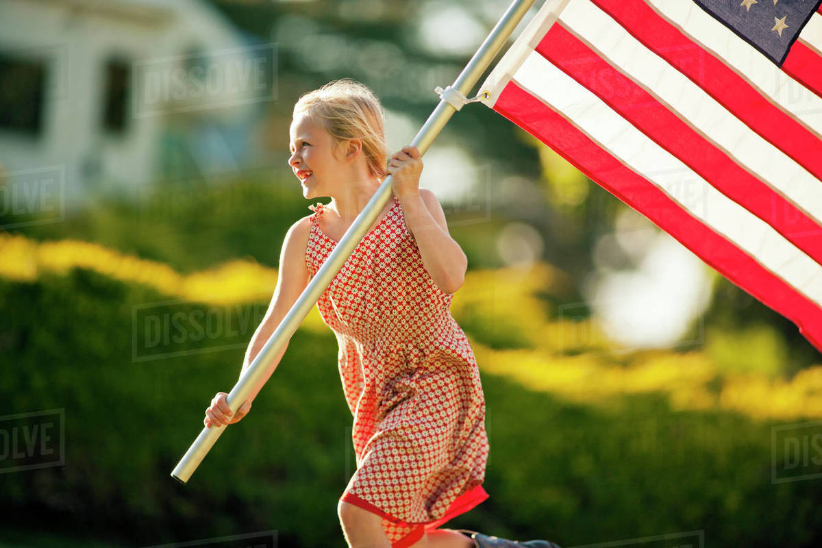 Smiling young girl carrying an American flag while running through her ...