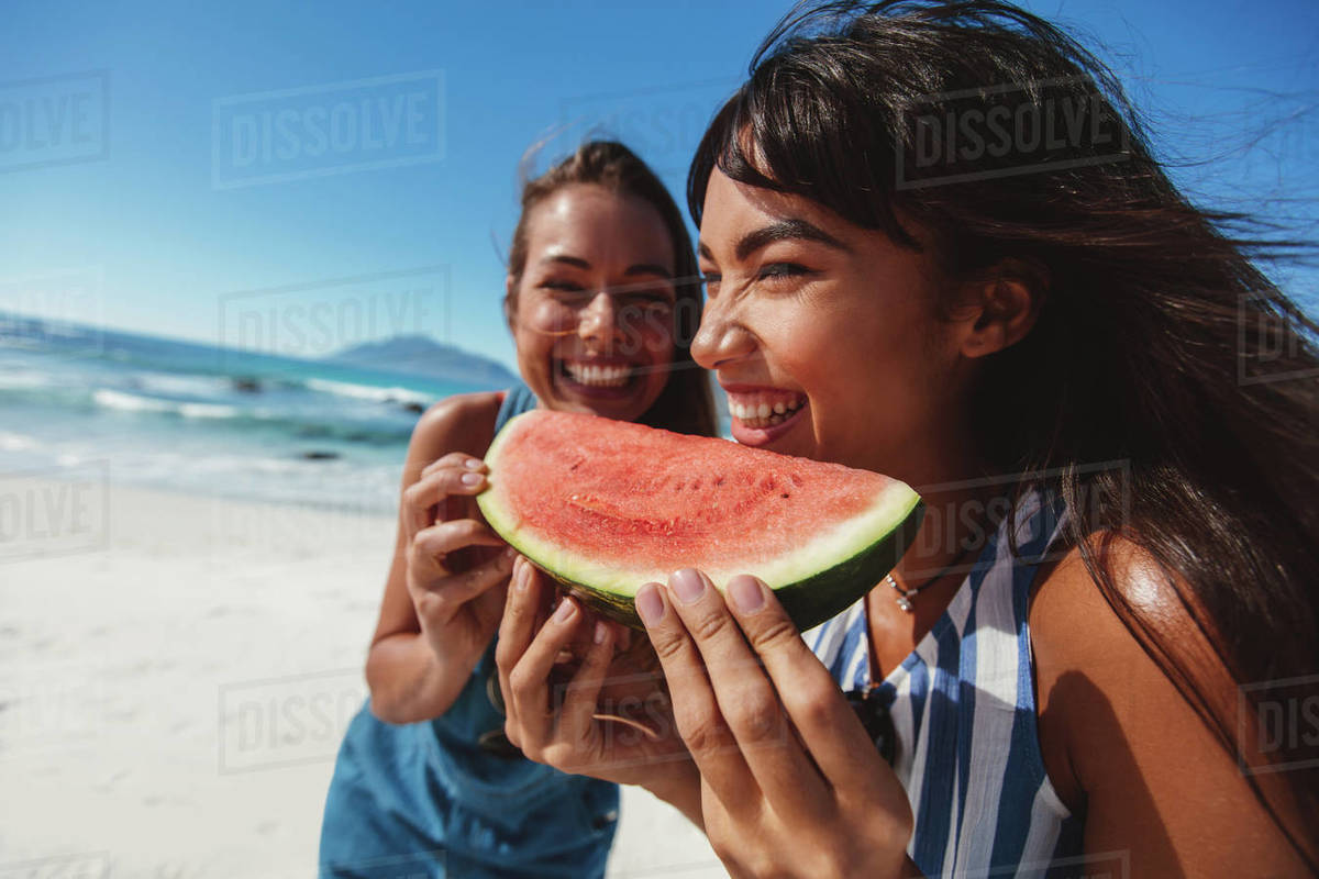 Two young women holding slice of watermelon and smiling on the beach ...