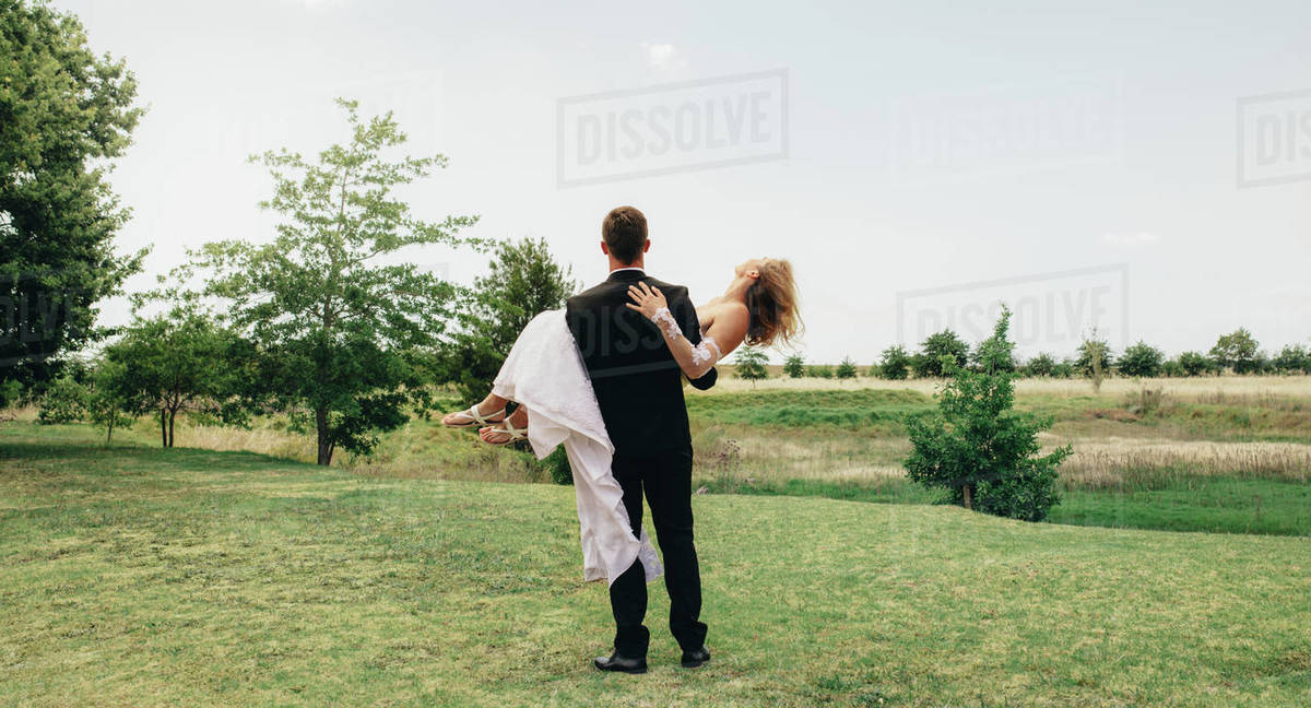 Bridegroom carrying his bride in his arms and standing outside. Rear ...