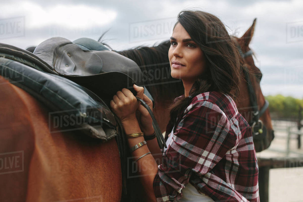 Beautiful young woman fixing a saddle on her horse. Cowgirl getting ...