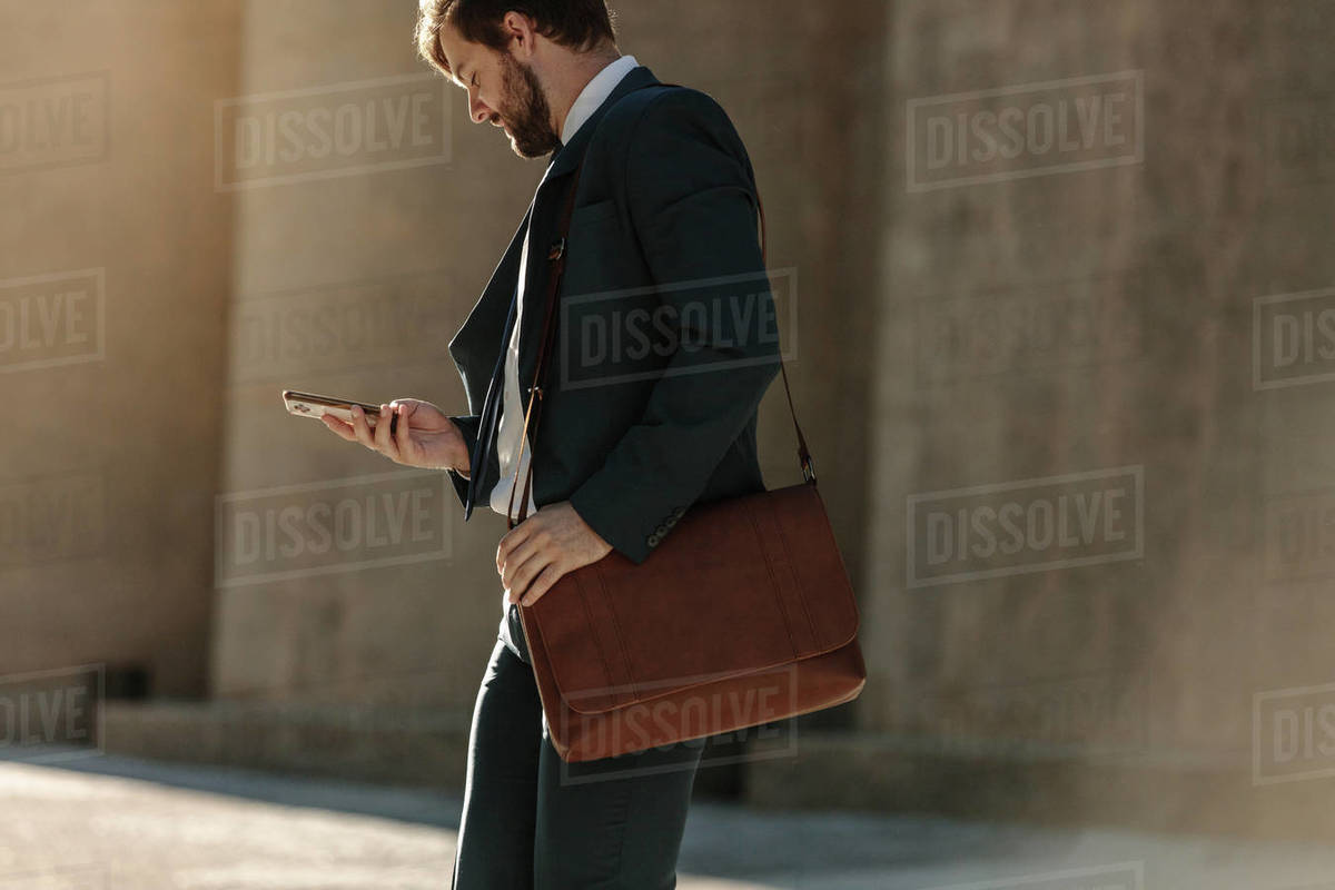 Businessman looking at his mobile phone while walking on street to ...