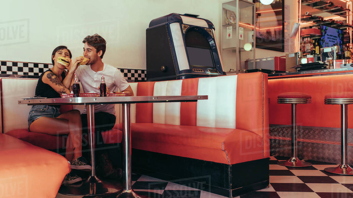 Couple sitting at a restaurant and eating burgers interlocking their ...
