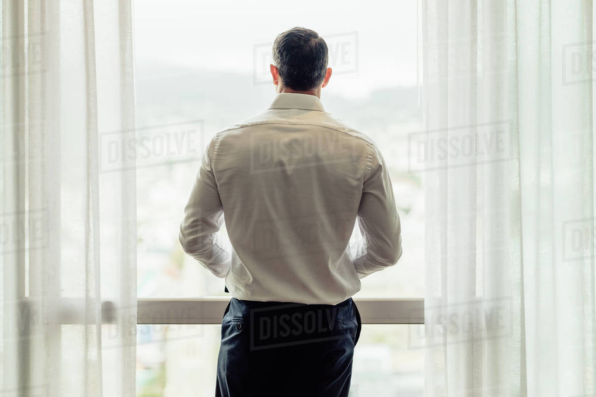 Rear view of man in formal wear standing at hotel room window and ...