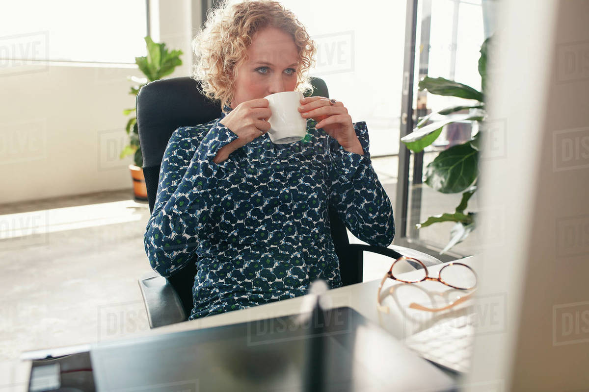 Portrait of young woman sitting at her desk drinking coffee and looking ...