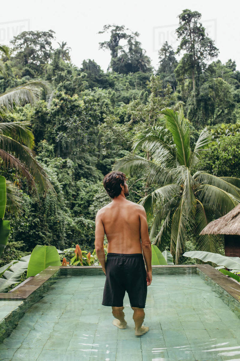 Rear view shot of young man standing in pool and looking away at a view ...