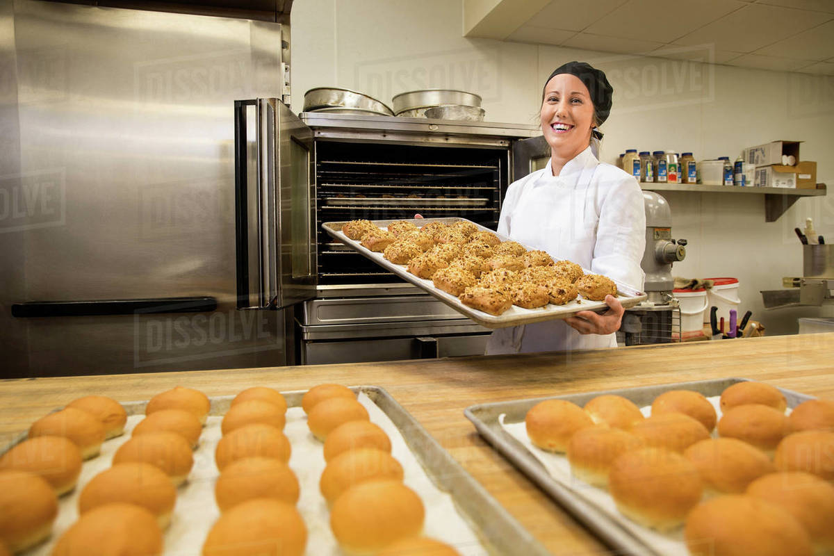 Female baker smiling in kitchen holding tray of rolls fresh out of the ...