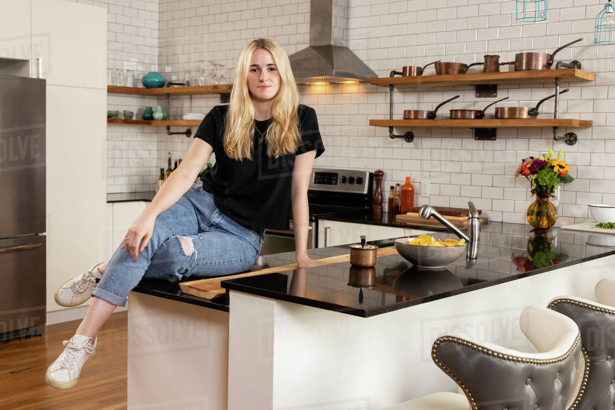 Young woman sitting on counter top in kitchen looking at camera with ...