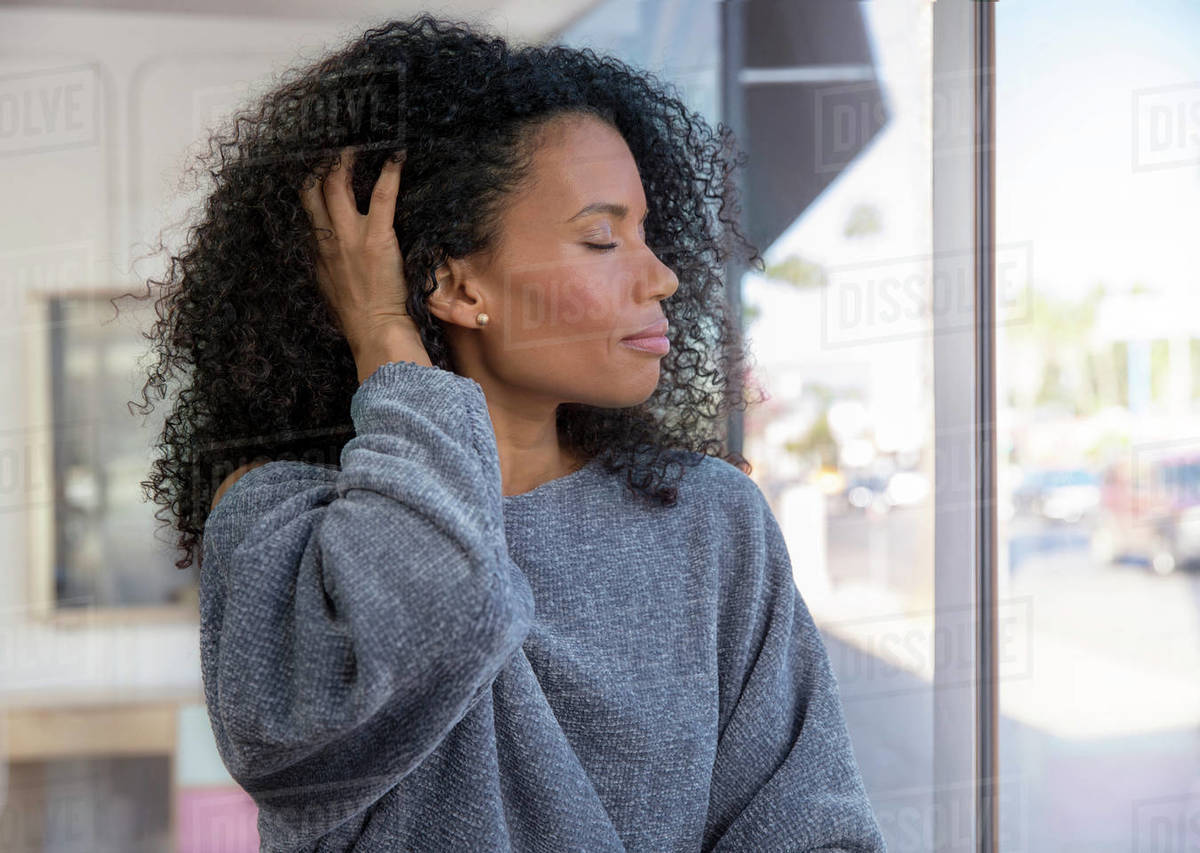 Mixed race, middle-aged woman runs her hands through her hair with her eyes closed looking out a ...