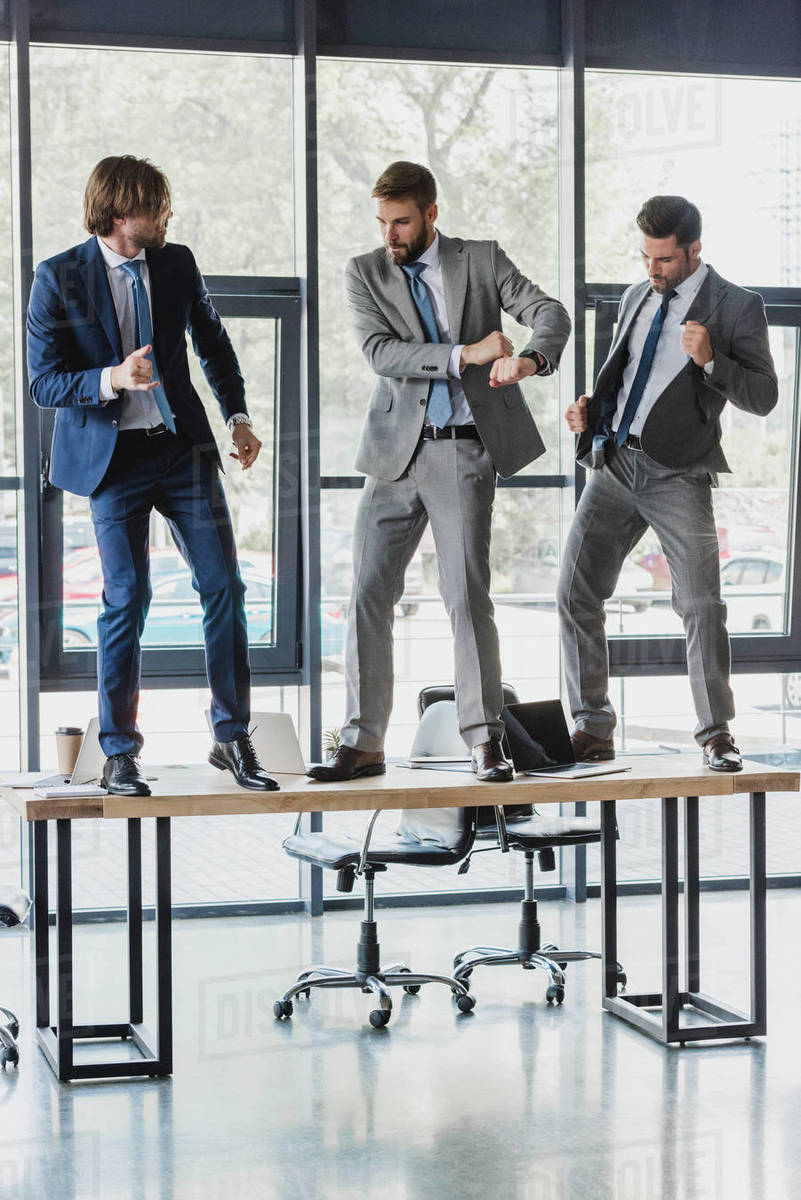 Three smiling young businessmen dancing on table in office - Royalty ...