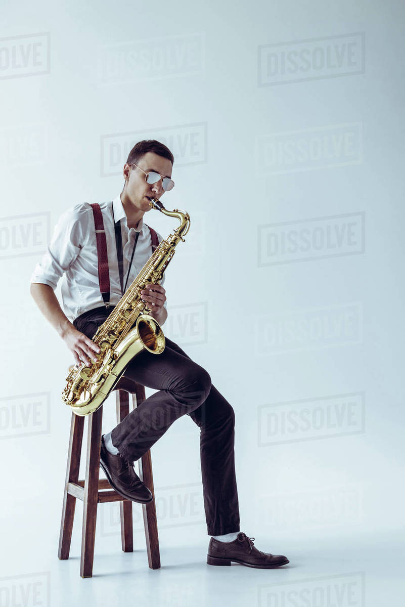 Handsome young performer sitting on stool and playing saxophone on grey ...