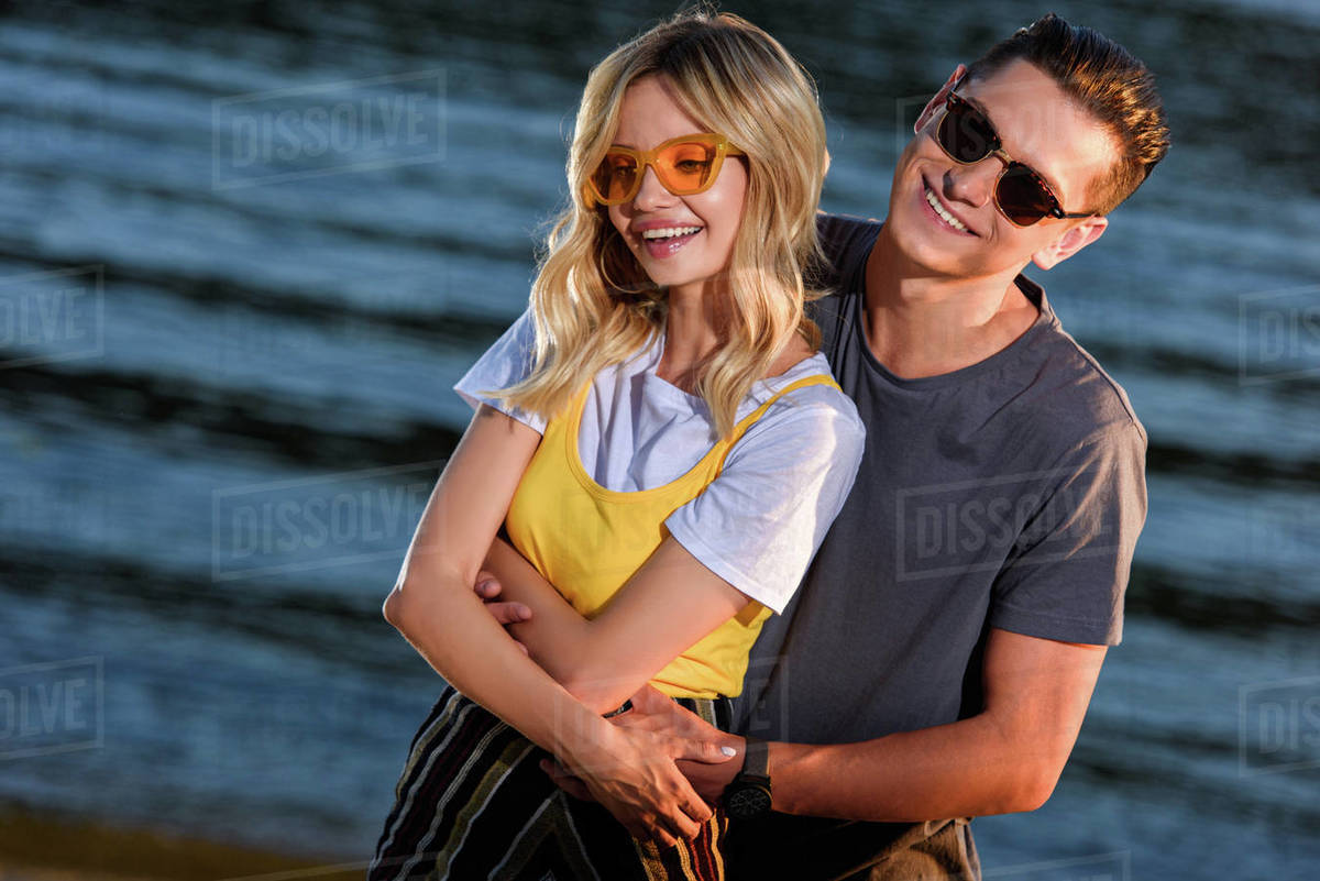 cheerful boyfriend cuddling girlfriend on river beach in evening ...
