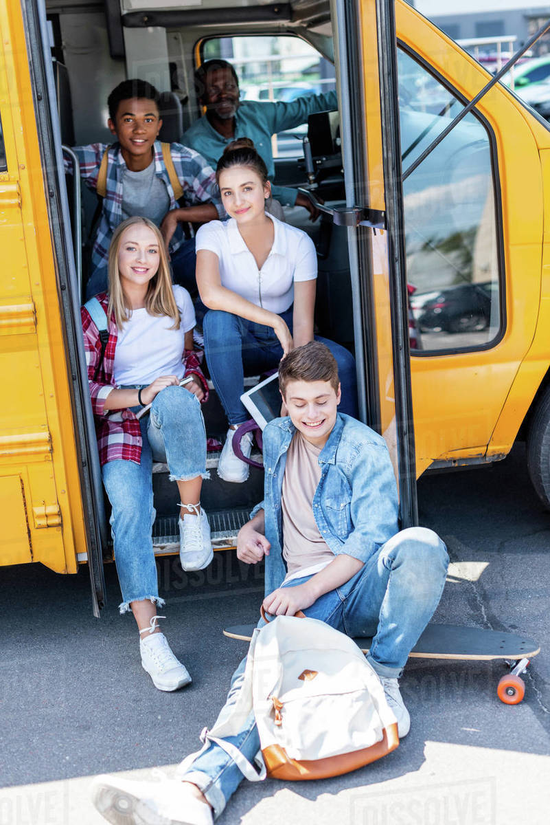 group of happy teen scholars sitting at school bus with driver inside ...