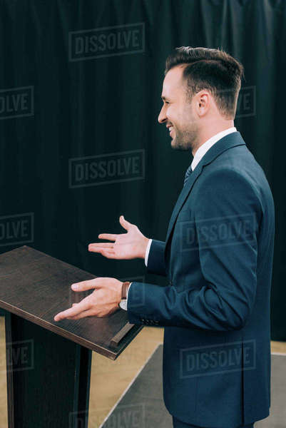 Side view of handsome lecturer standing and gesturing at podium tribune ...