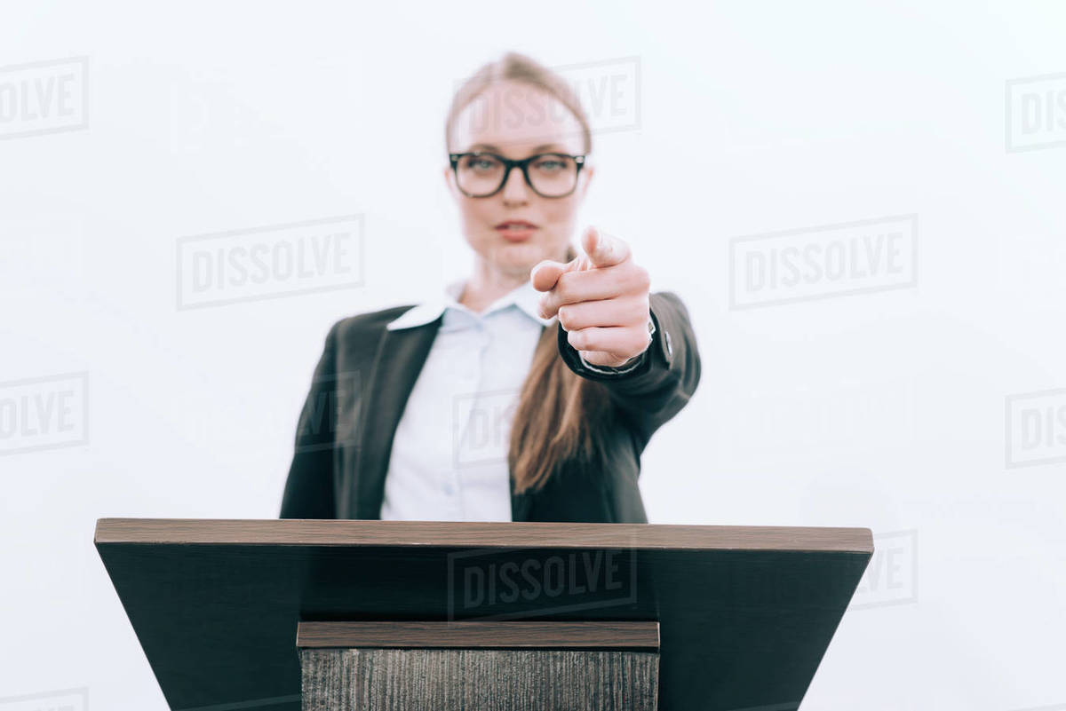 Selective focus of attractive lecturer pointing on camera from podium ...