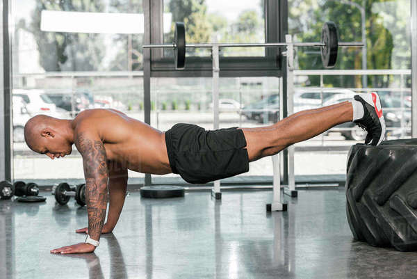 side view of muscular african american man doing push ups with tyre in ...