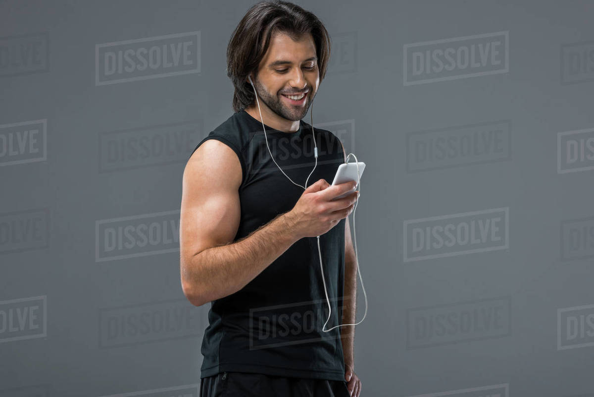 Smiling muscular man in earphones using smartphone isolated on grey ...