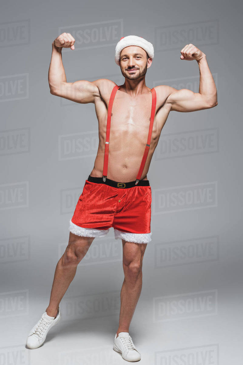 Young muscular man in christmas hat showing muscles isolated on grey ...