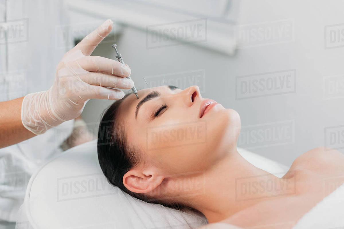 Partial view of cosmetologist putting needle on womans forehead during ...