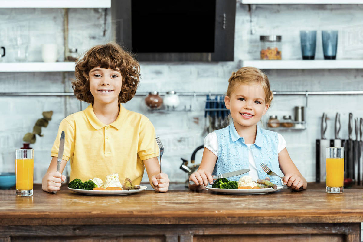 Adorable kids eating vegetables and smiling at camera in kitchen ...