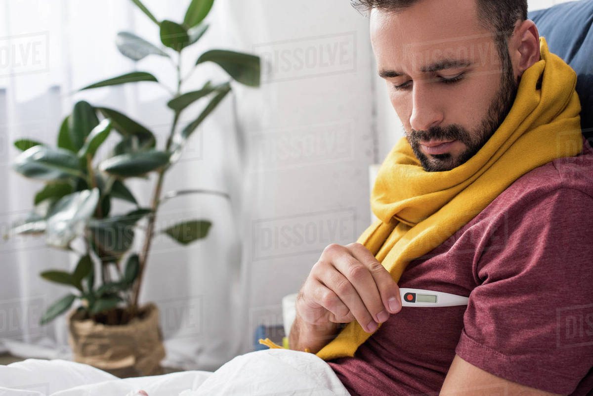 Sick young man measuring temperature with electronic thermometer in bed ...
