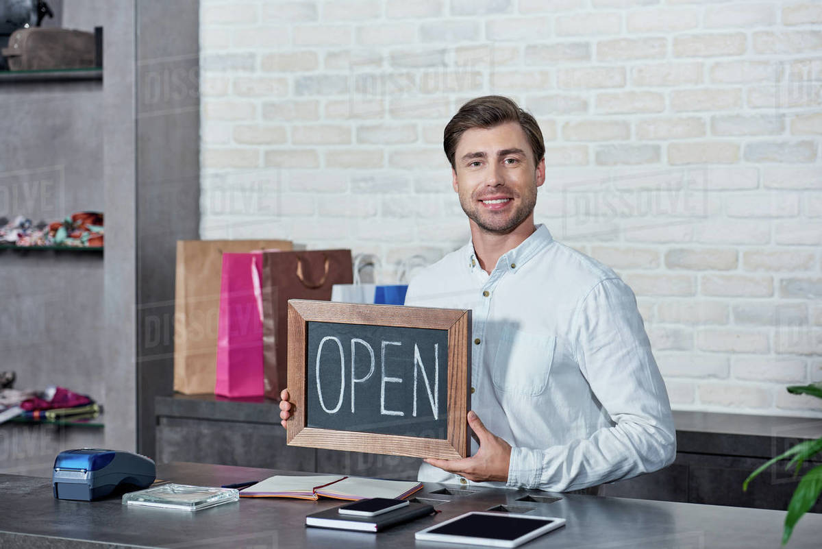 Handsome young salesman holding sign open and smiling at camera in shop ...