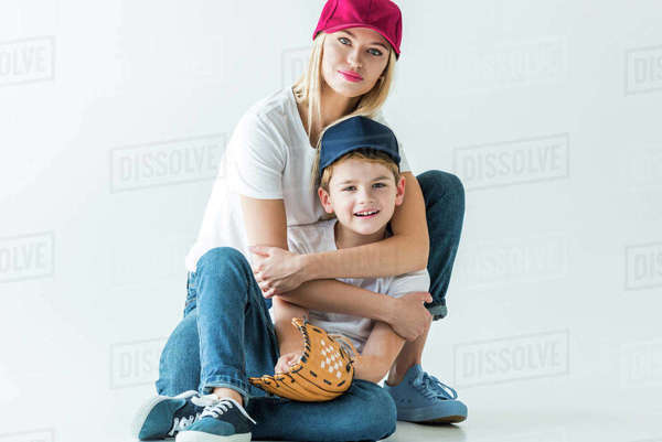 Smiling mother hugging son and sitting on floor with baseball glove on ...