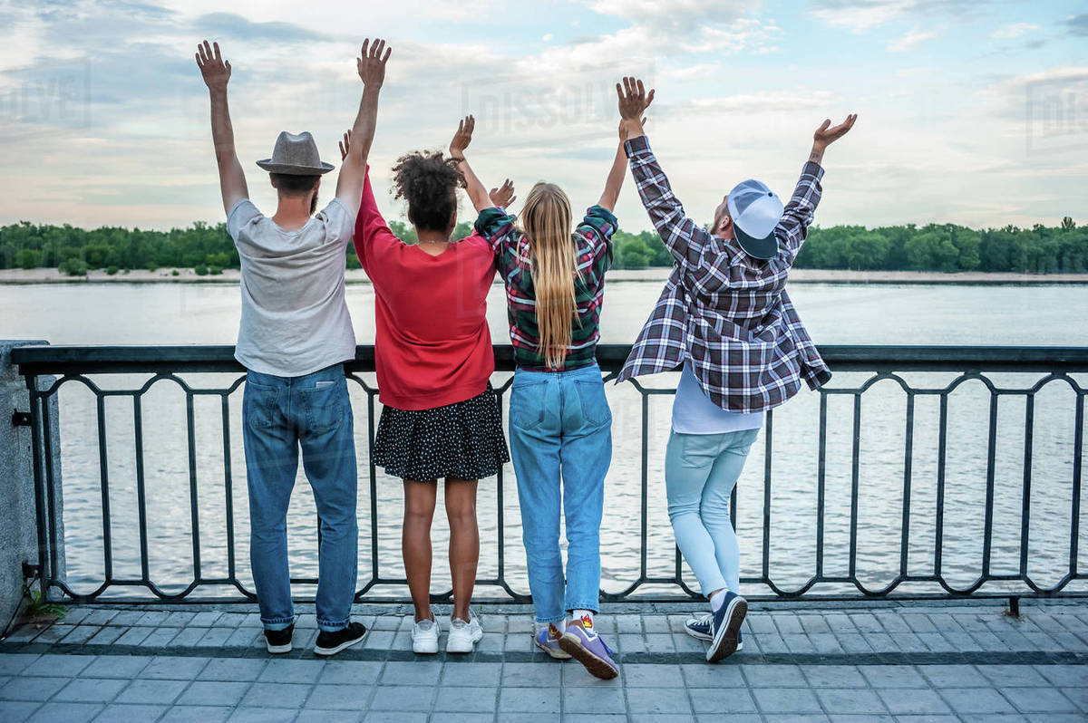 Back view of four multiethnic friends raising hands at riverbank ...