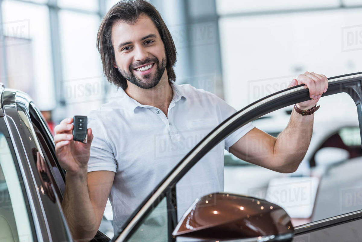 Portrait of happy man with car key standing at new car in dealership ...