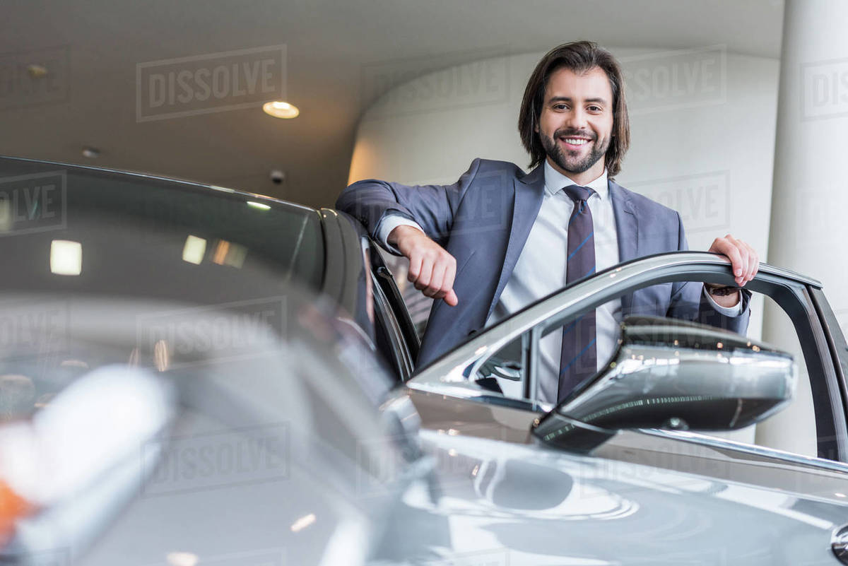 Happy stylish businessman standing at new car at dealership salon ...