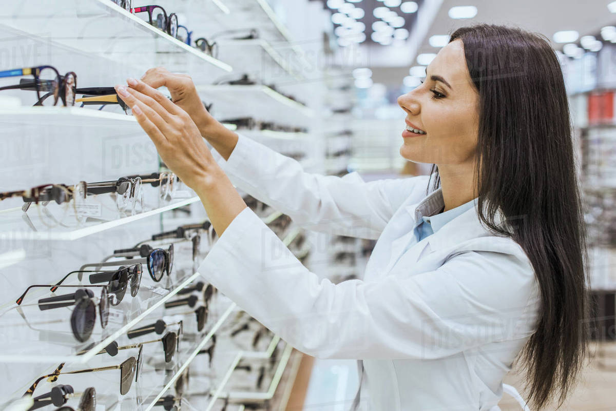 Beautiful smiling optician taking eyeglasses from shelves in ophthalmic ...