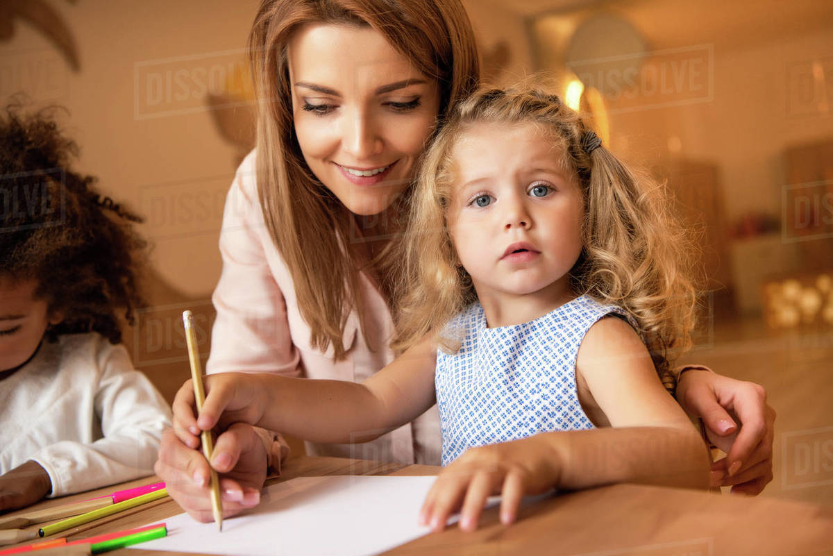 Cheerful educator helping adorable kid drawing in kindergarten ...