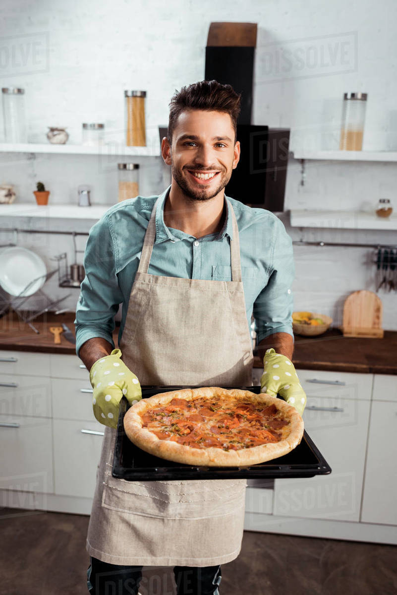 High angle view of smiling young man in apron and potholders holding ...